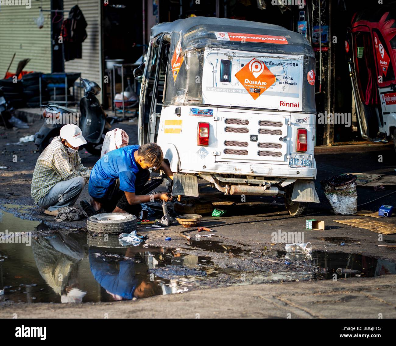 A mechanic repairs a three-wheeled auto rickshaw on a busy street. A ...