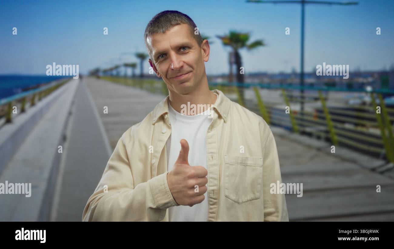 Smiling man gives thumbs-up on seaside promenade with ocean and palm trees in background ...