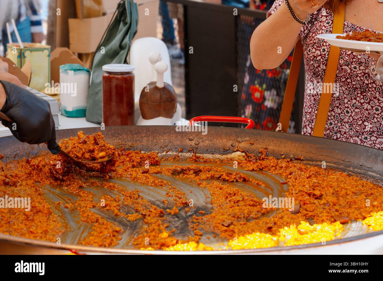 A food vendor scoops hot paella from a wide pan as a woman holds her ...