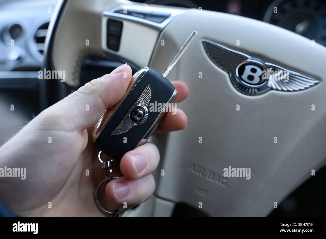 This image features a hand holding a Bentley key fob inside a luxury vehicle, with the high-end ...
