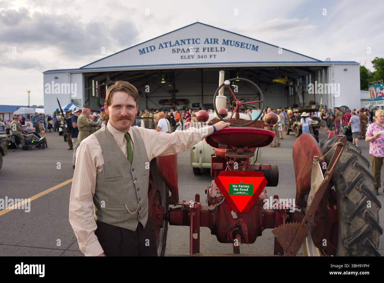 A man in period attire stands proudly beside a vintage red tractor at ...