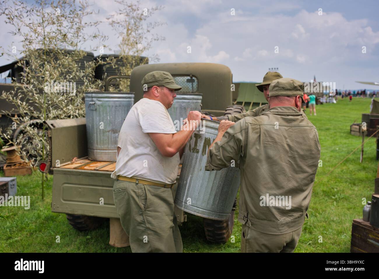 Two men in WWII-era military uniforms lift metal garbage cans onto the ...