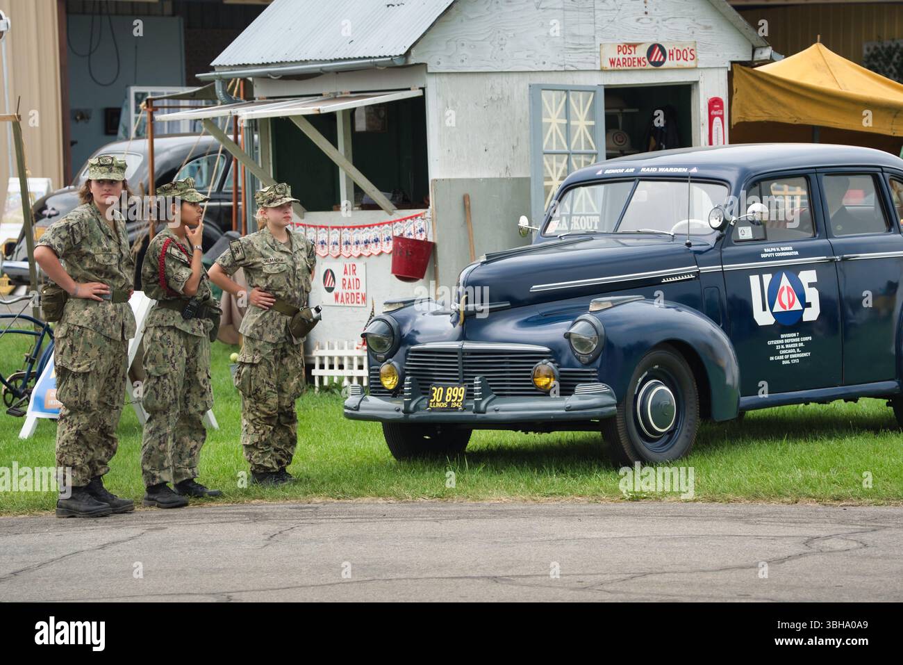 Three women in military-style camouflage uniforms stand near a vintage ...