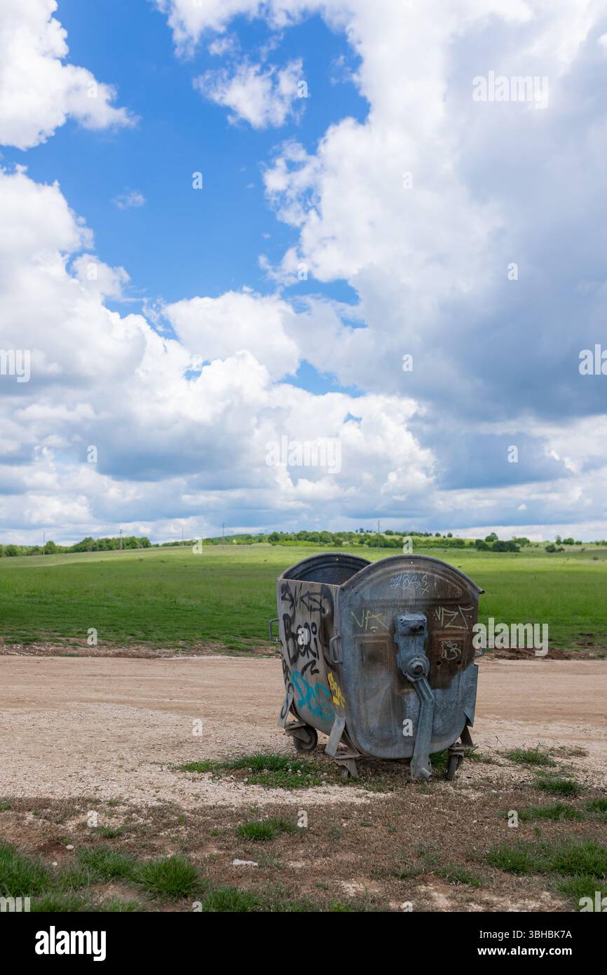 Graffitied garbage bin stands by rural road with green pastures ...
