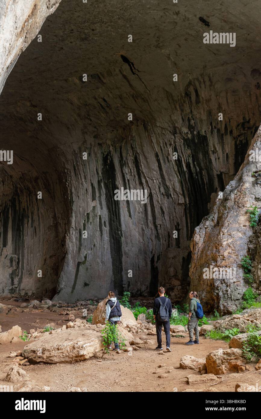 Group of hikers stands at massive cave entrance with towering vertical ...
