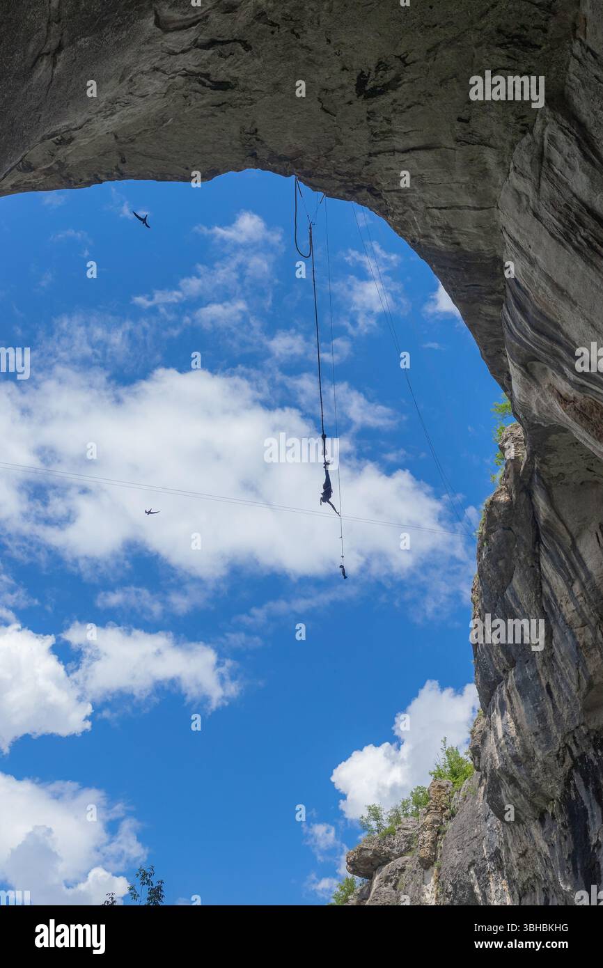 Person hanging mid-air on rope under vast cave arch and blue sky ...