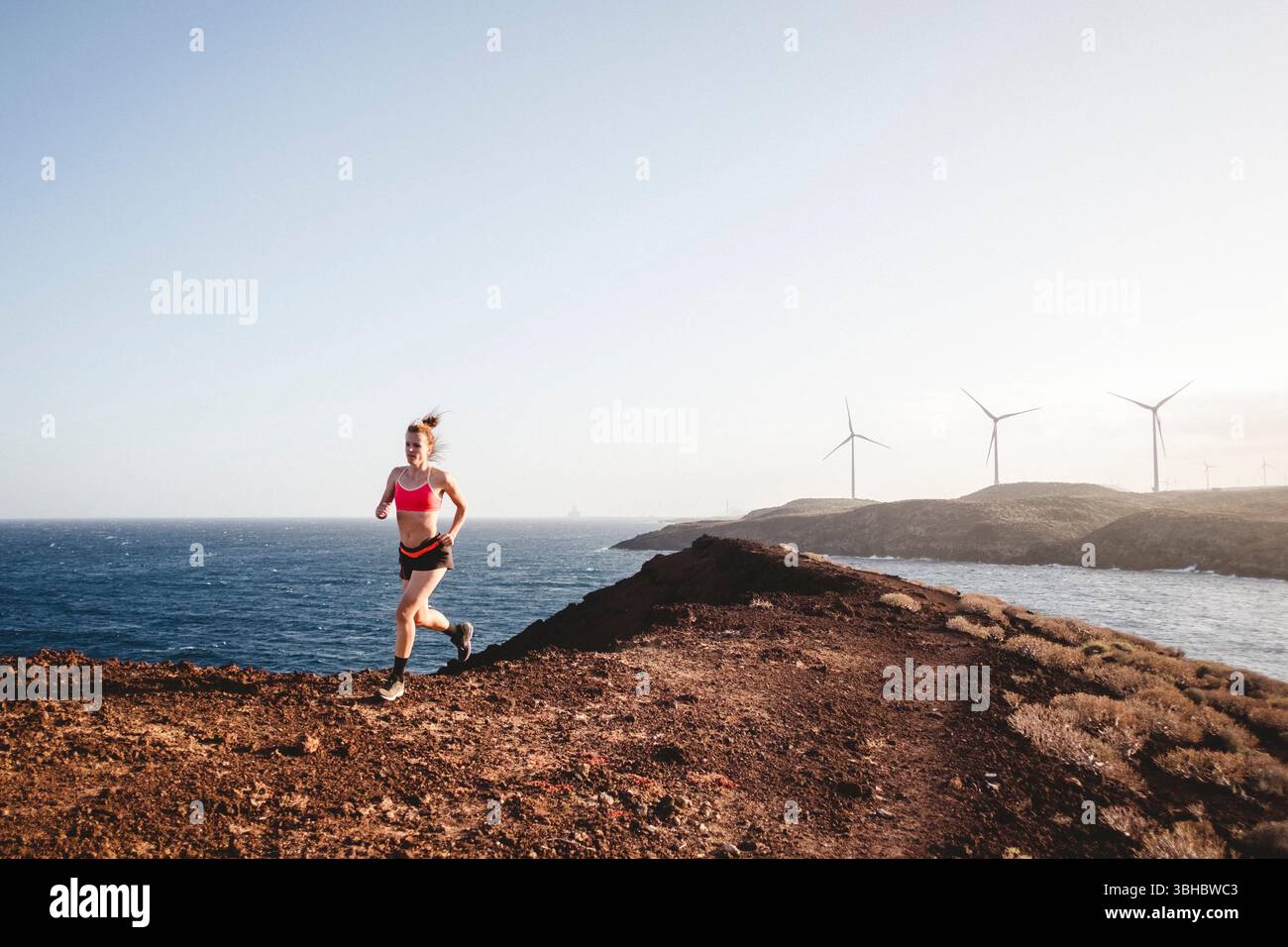 Female runner full body on trail by sea with wind mills in background ...