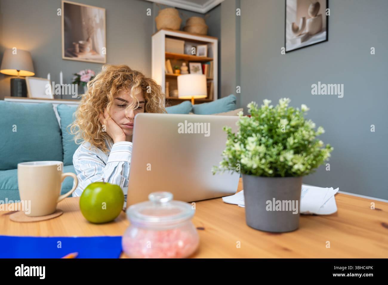 Young woman struggling with focus while working on laptop on floor, surrounded by printed ...