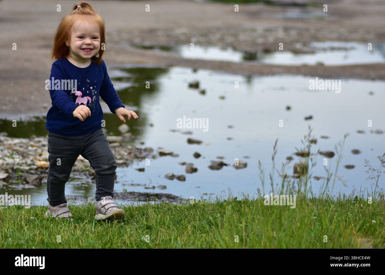 Joyful toddler girl with red hair walks near a puddle outdoors, making ...