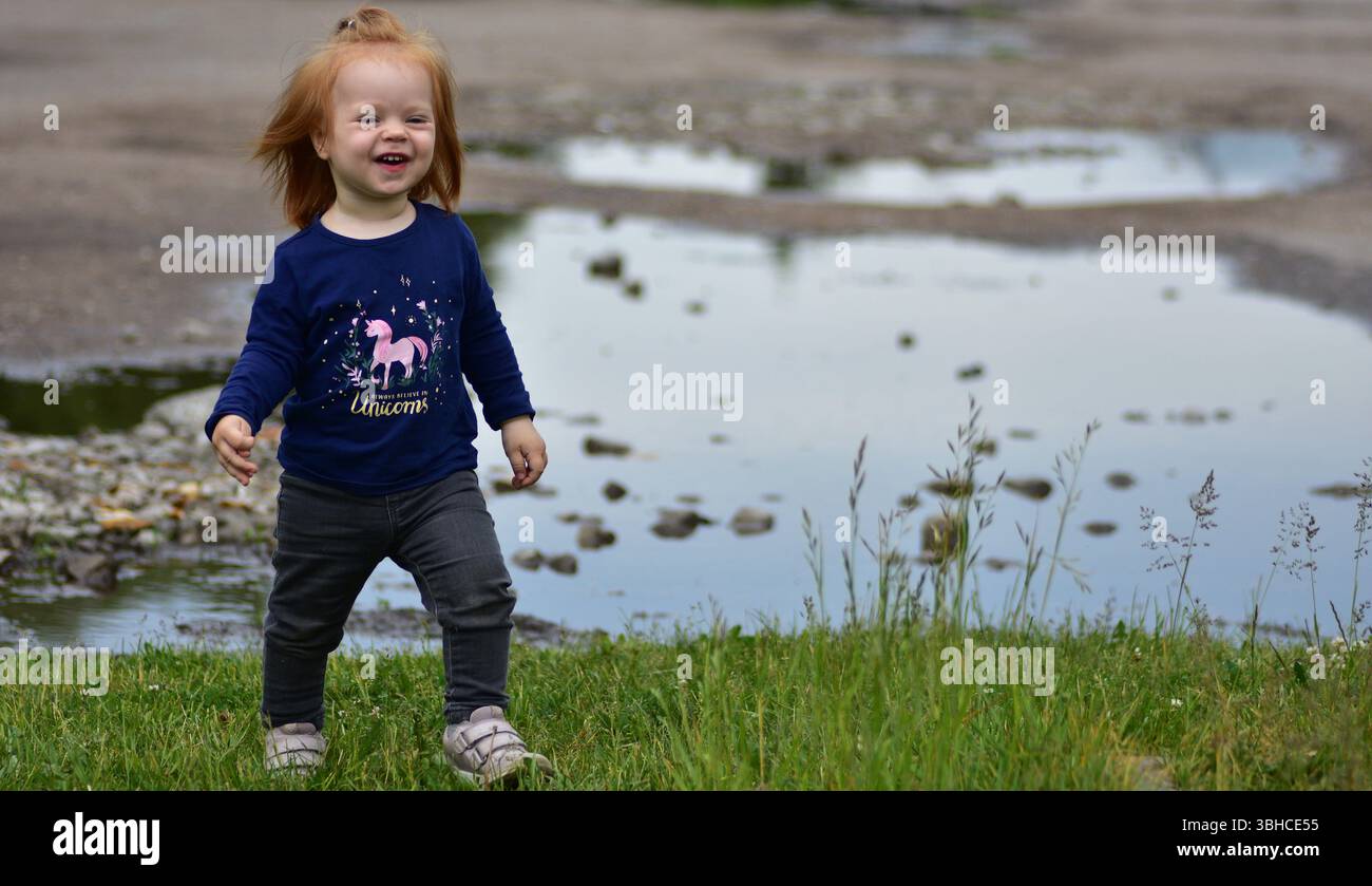 Joyful toddler girl with red hair walks near a puddle outdoors, making ...