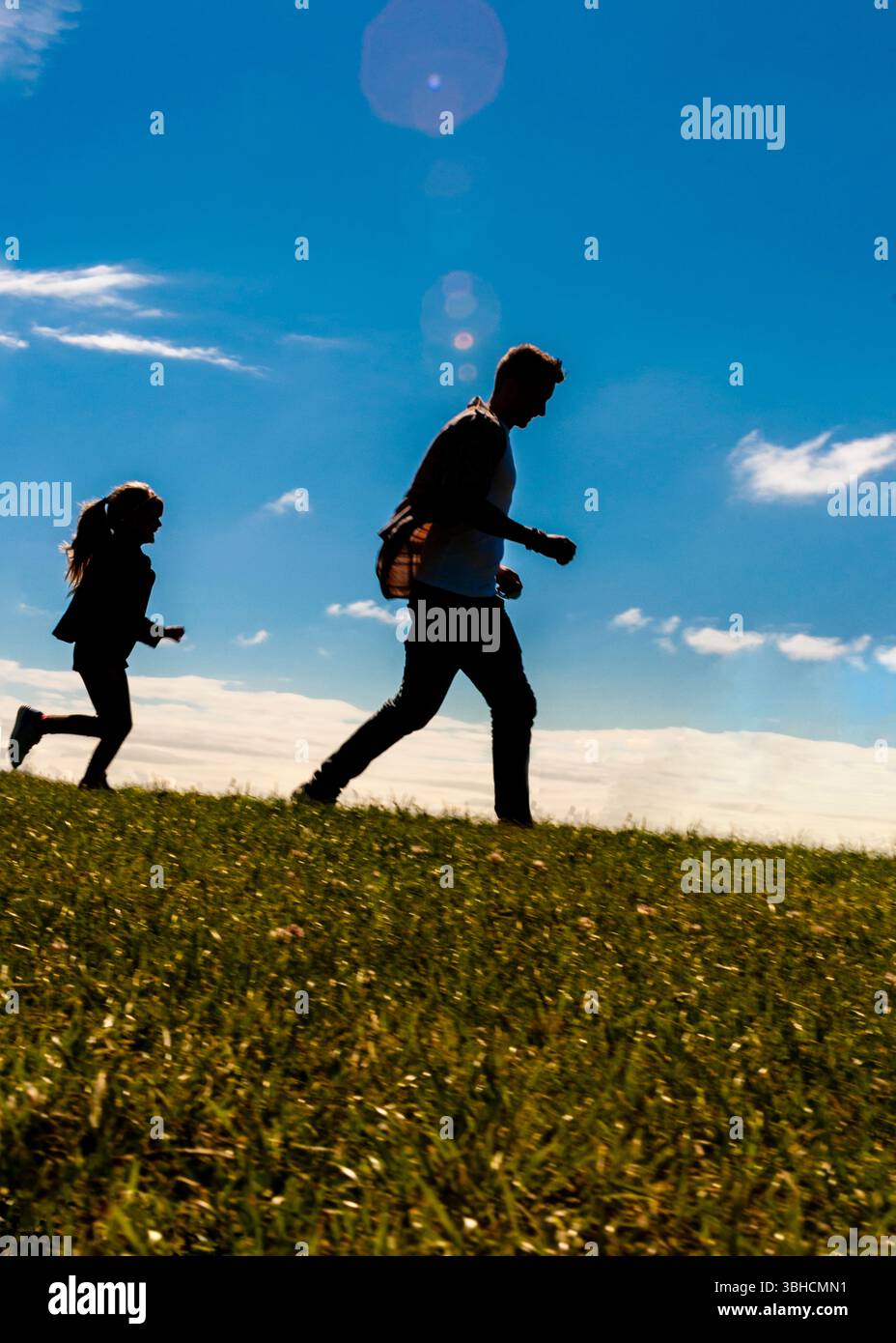 A father and daughter run across a grassy hill under a bright blue sky ...