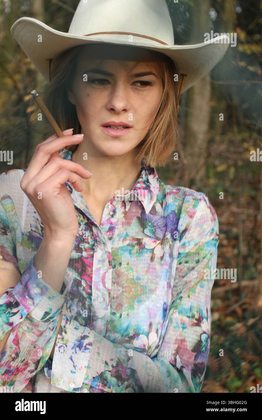 Attractive young woman in cowboy hat with thin cigar or cigarillos looking away from camera ...