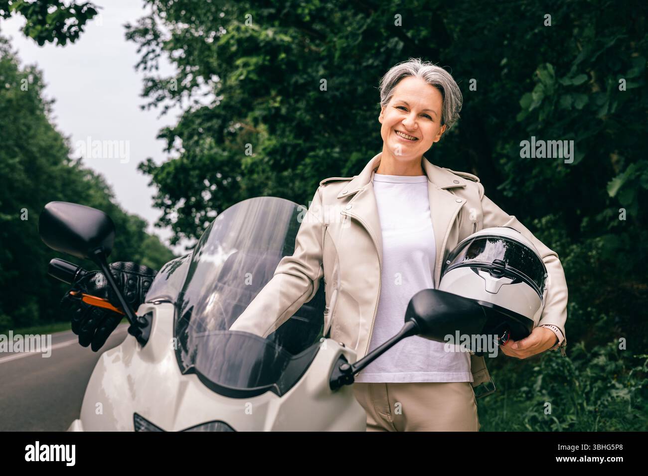 Smiling gray-haired senior woman in beige outfit holding motorcycle ...
