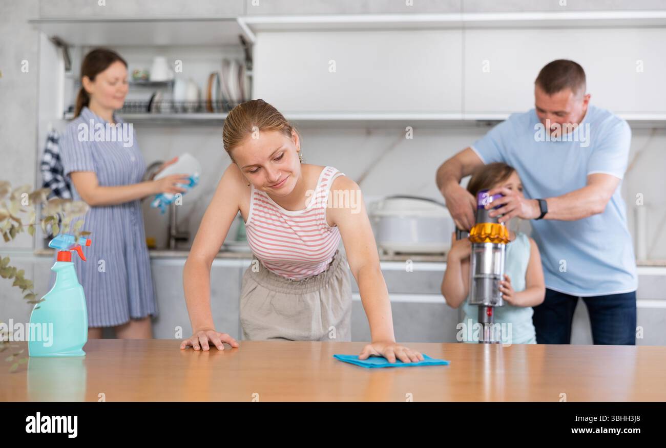 Teen girl cleans table with rag, family clean kitchen in background ...