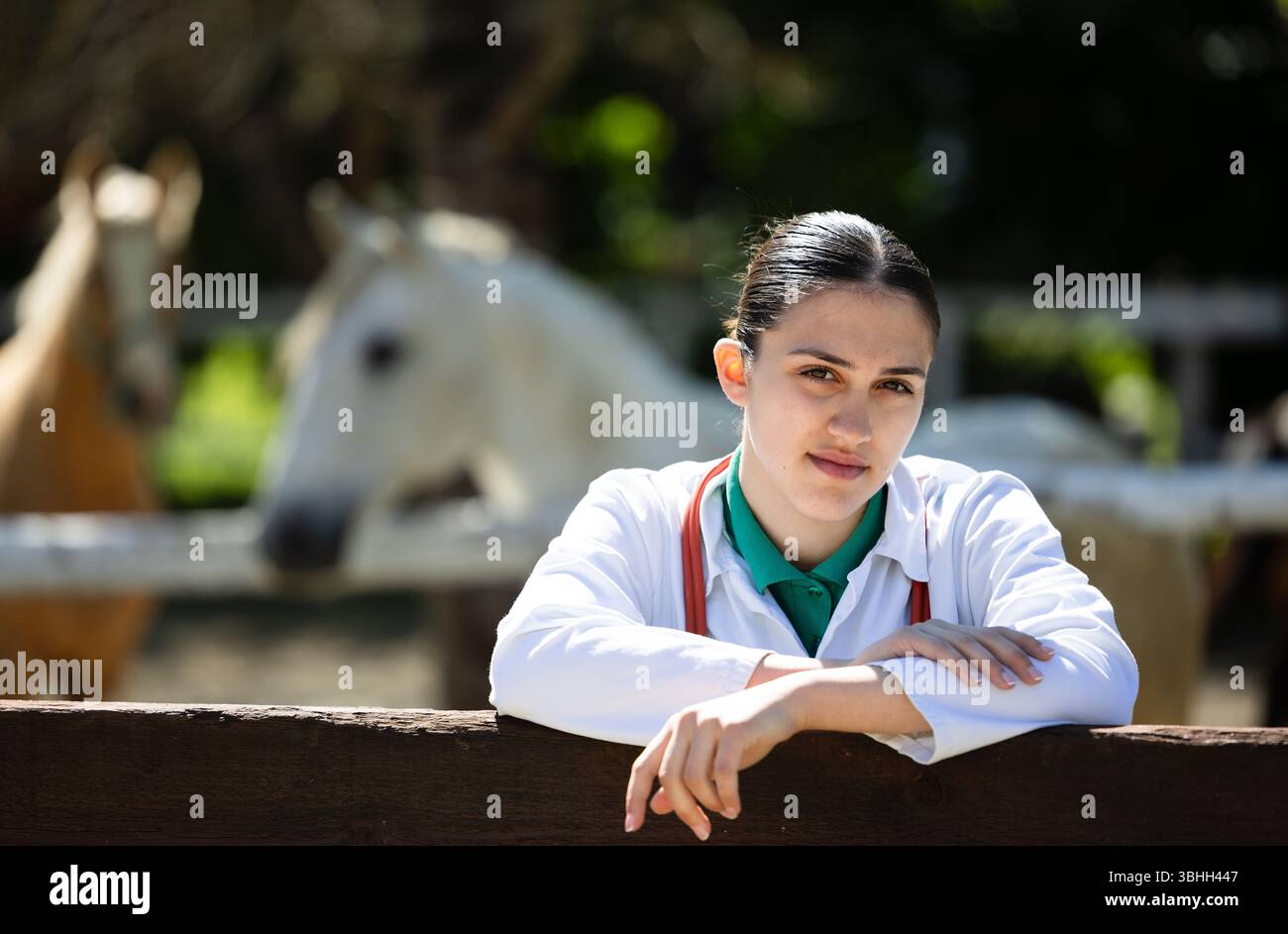 Portrait of female veterinarian stands by wooden fence in sunny stable ...