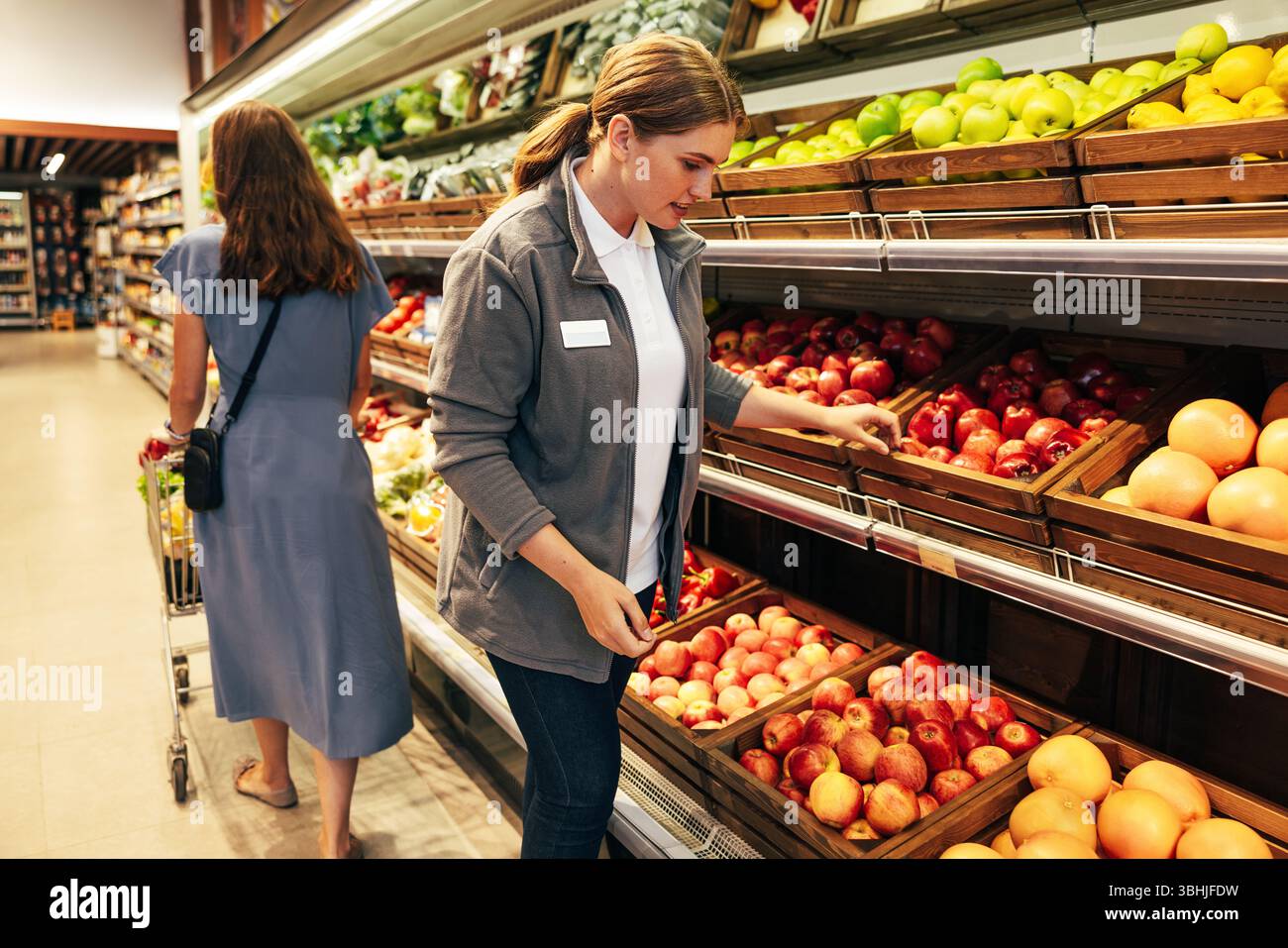 Female grocery store worker stands by the shelves as buyer with a cart ...