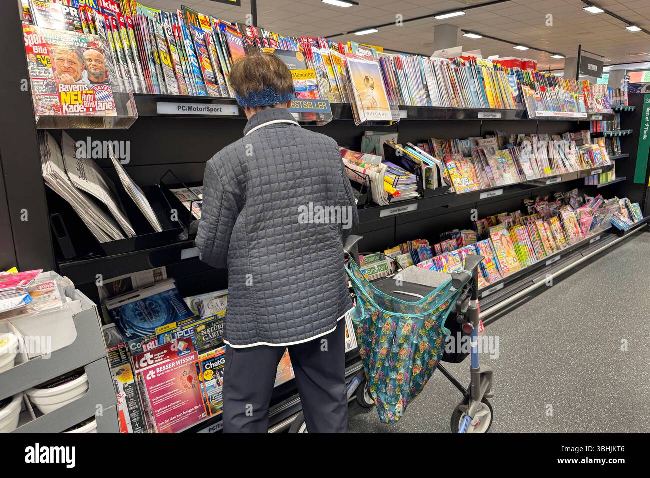 Alte Frau mit Rollator steht vor einem Regal mit Zeitschriften und ...