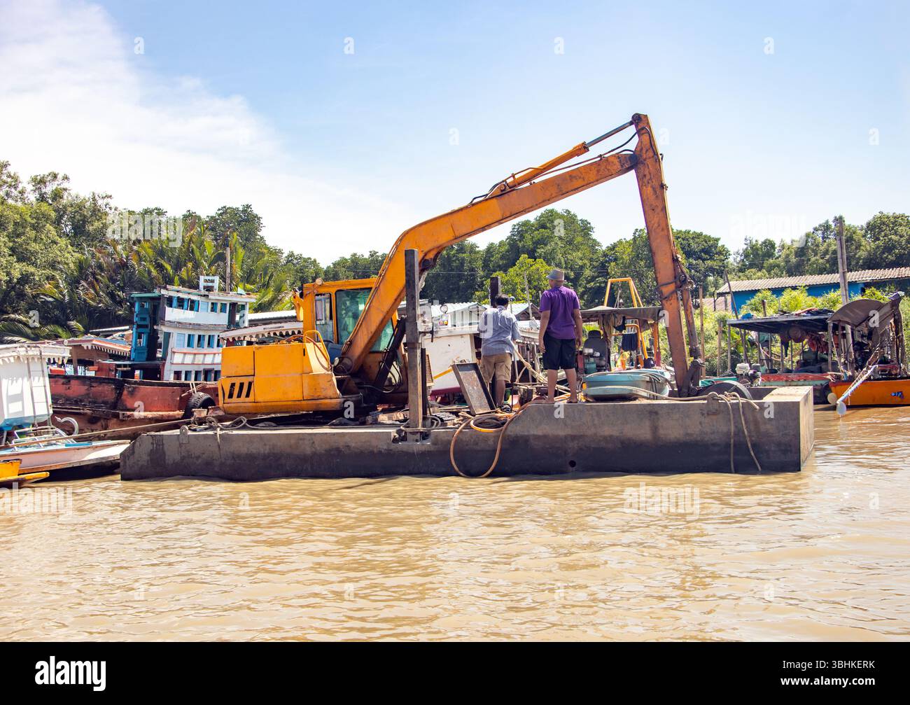 An excavator with workers on a floating barge at a riverside ...