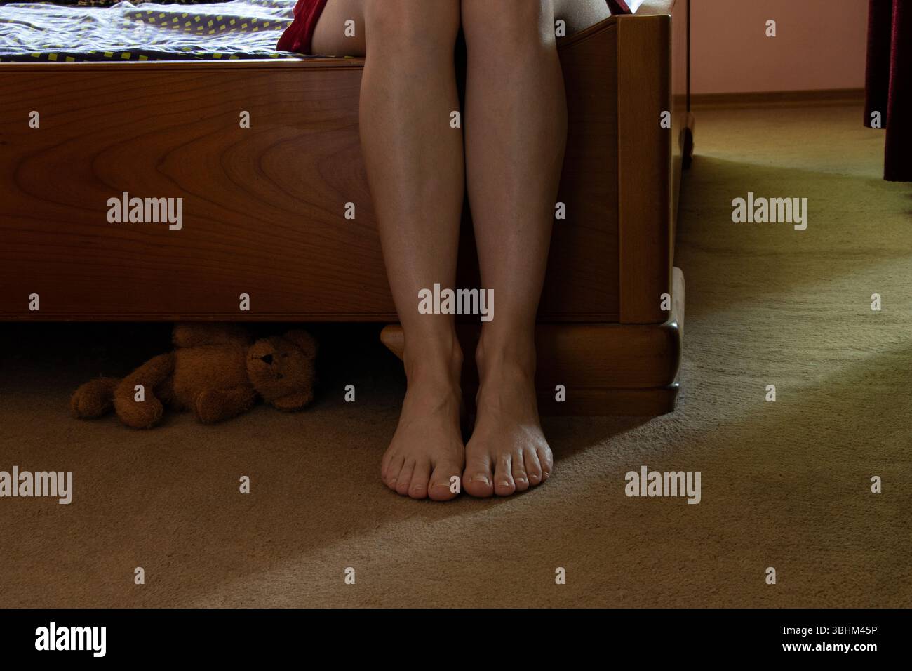 Legs of a girl sitting on a bed and a teddy bear on the floor in the ...