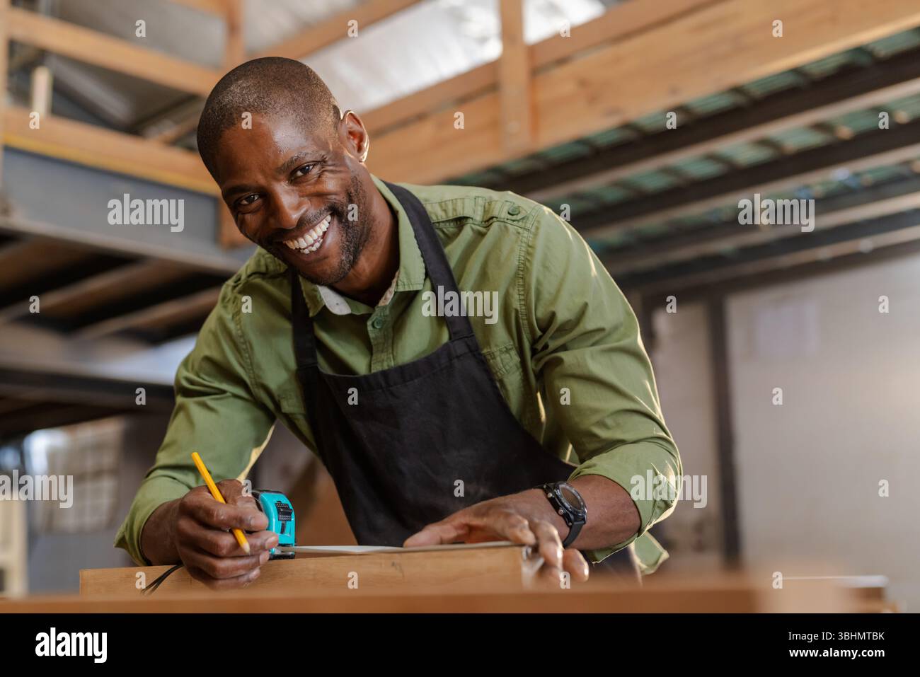 African American man in apron bending over bench marking wood plank ...