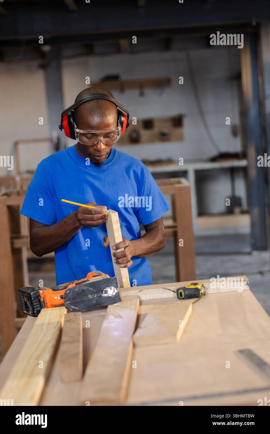 African American man wearing goggles and earmuffs marking timber with ...