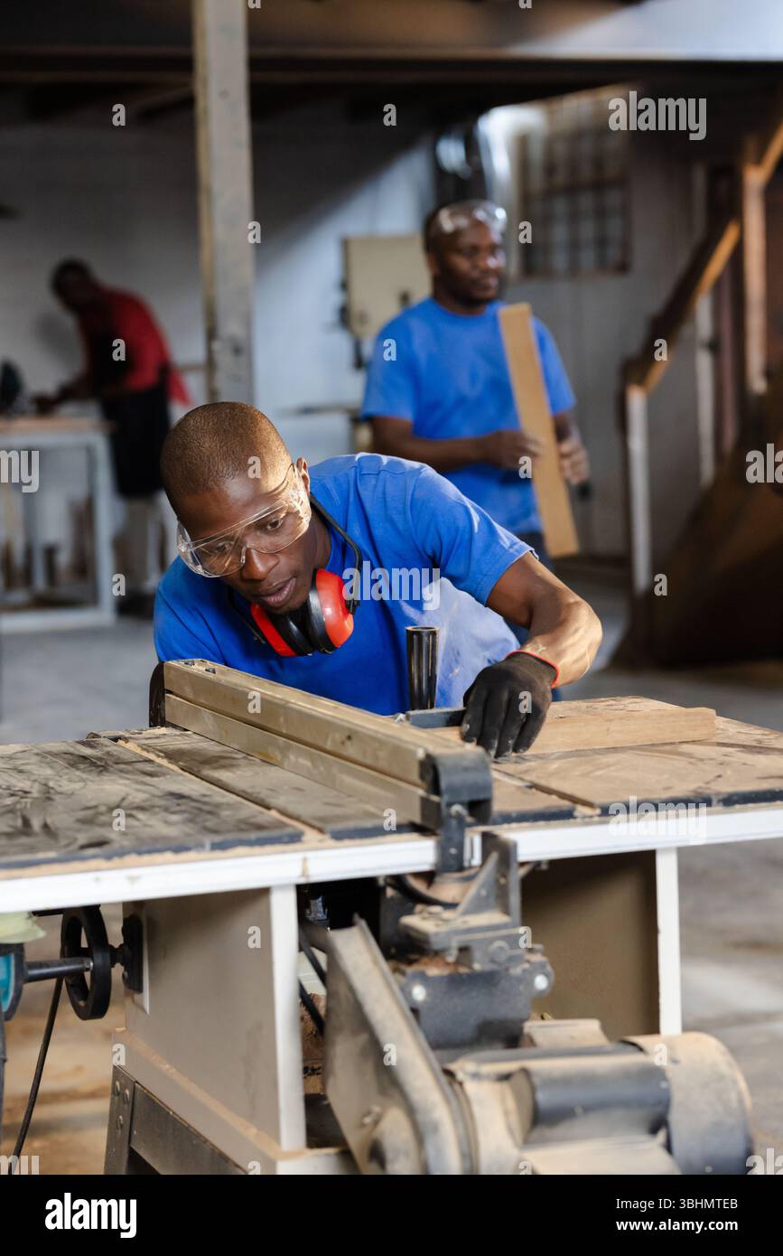 African American coworkers operating table saw handling planks with ...