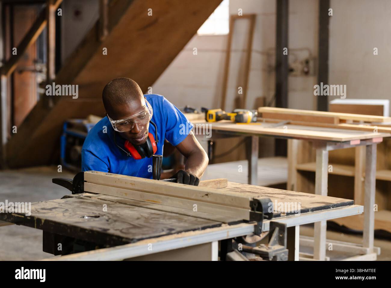 African American man wearing safety goggles guiding wooden plank ...