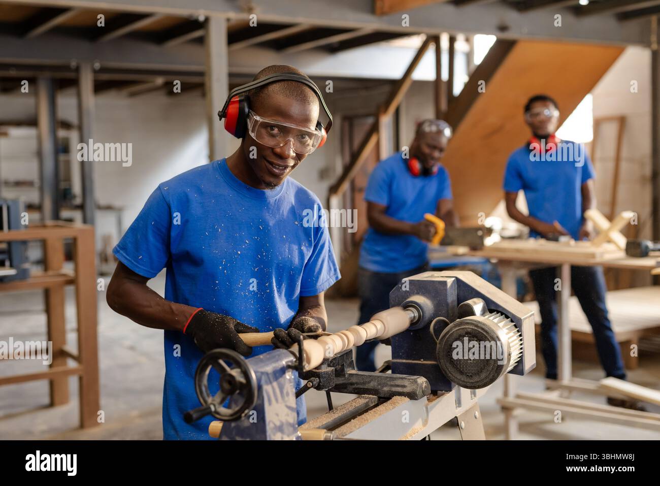 African American male coworkers operating wood lathe and sanding planks ...