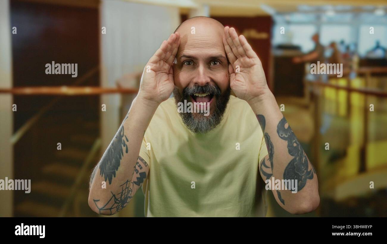 Bearded man with tattoos playfully covering face in a hotel lobby ...