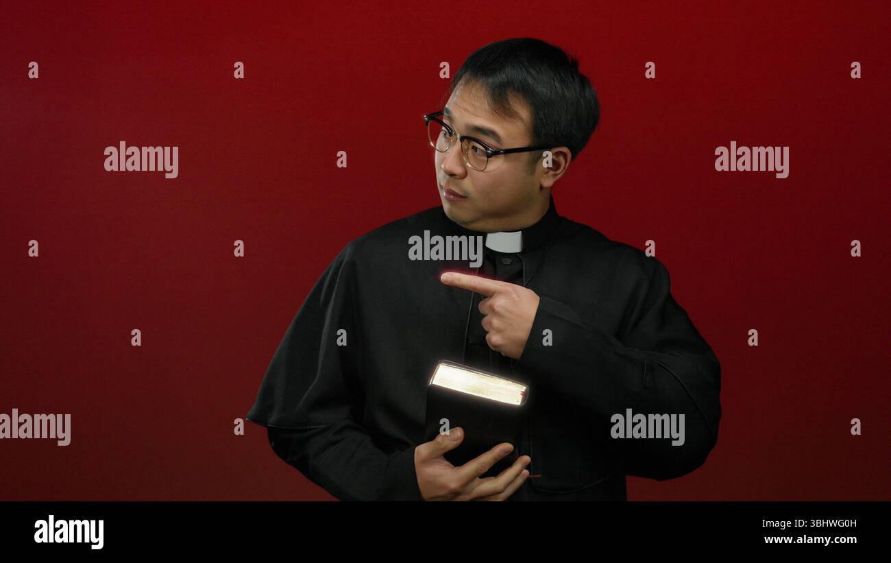 Asian priest in black robe holding bible and pointing on red background ...