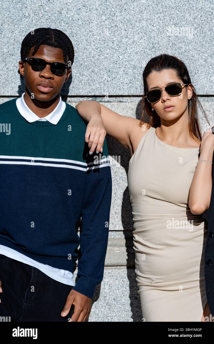 Two fashionable models wearing sunglasses pose against a gray wall ...