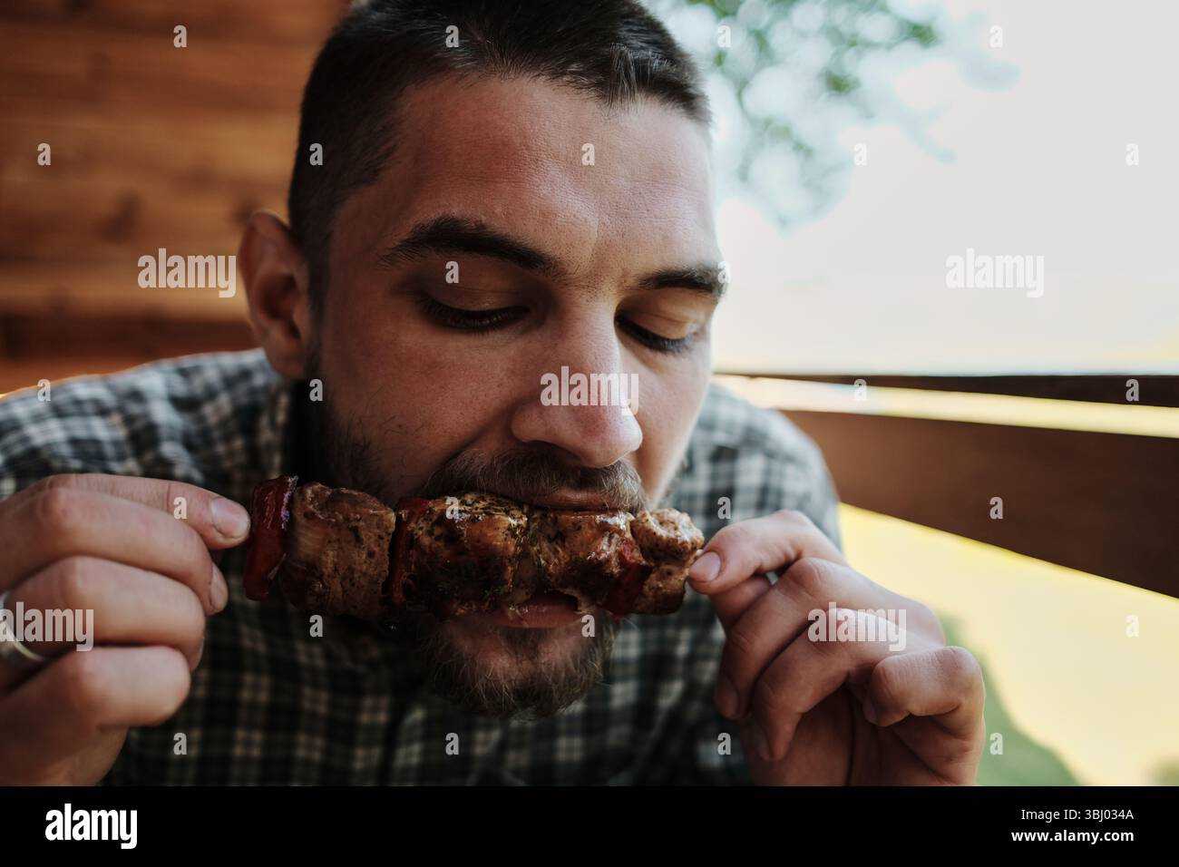 Man biting into a juicy grilled meat skewer, enjoying a rustic outdoor ...