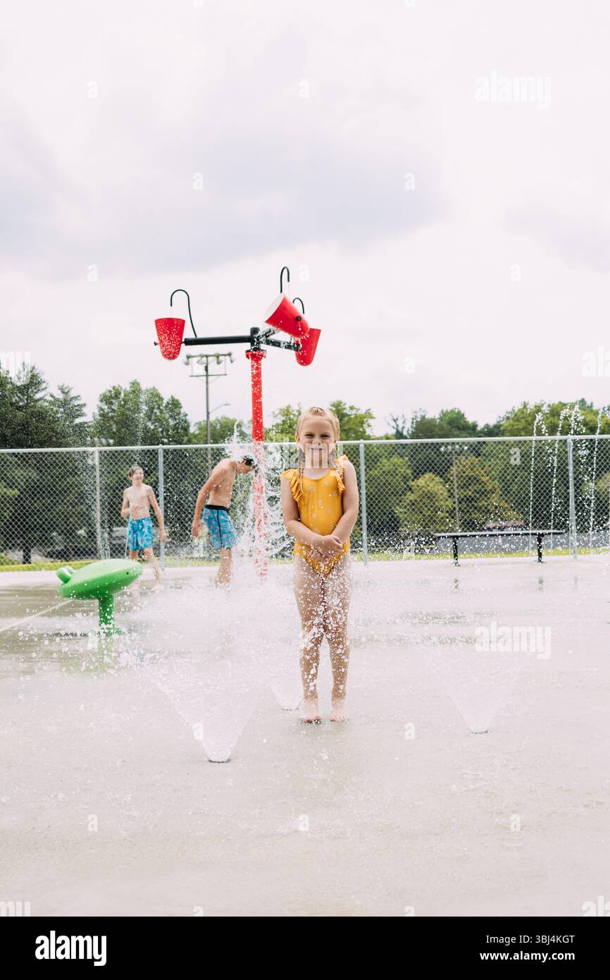 Smiling child standing in sprinklers while kids play in splash pad ...