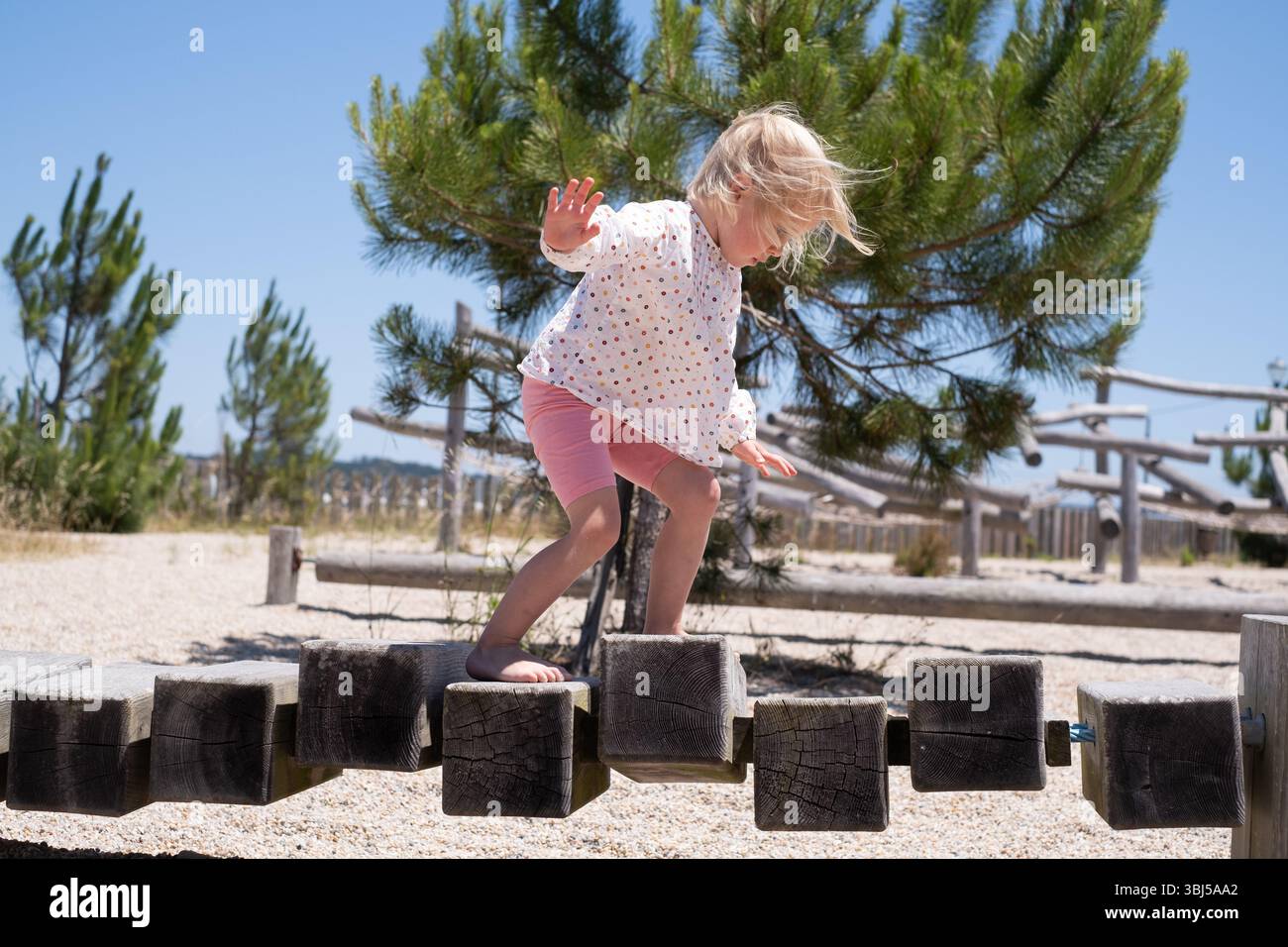 A 3-year-old blonde girl playing on a playground, trying to walk across a balance beam Stock ...