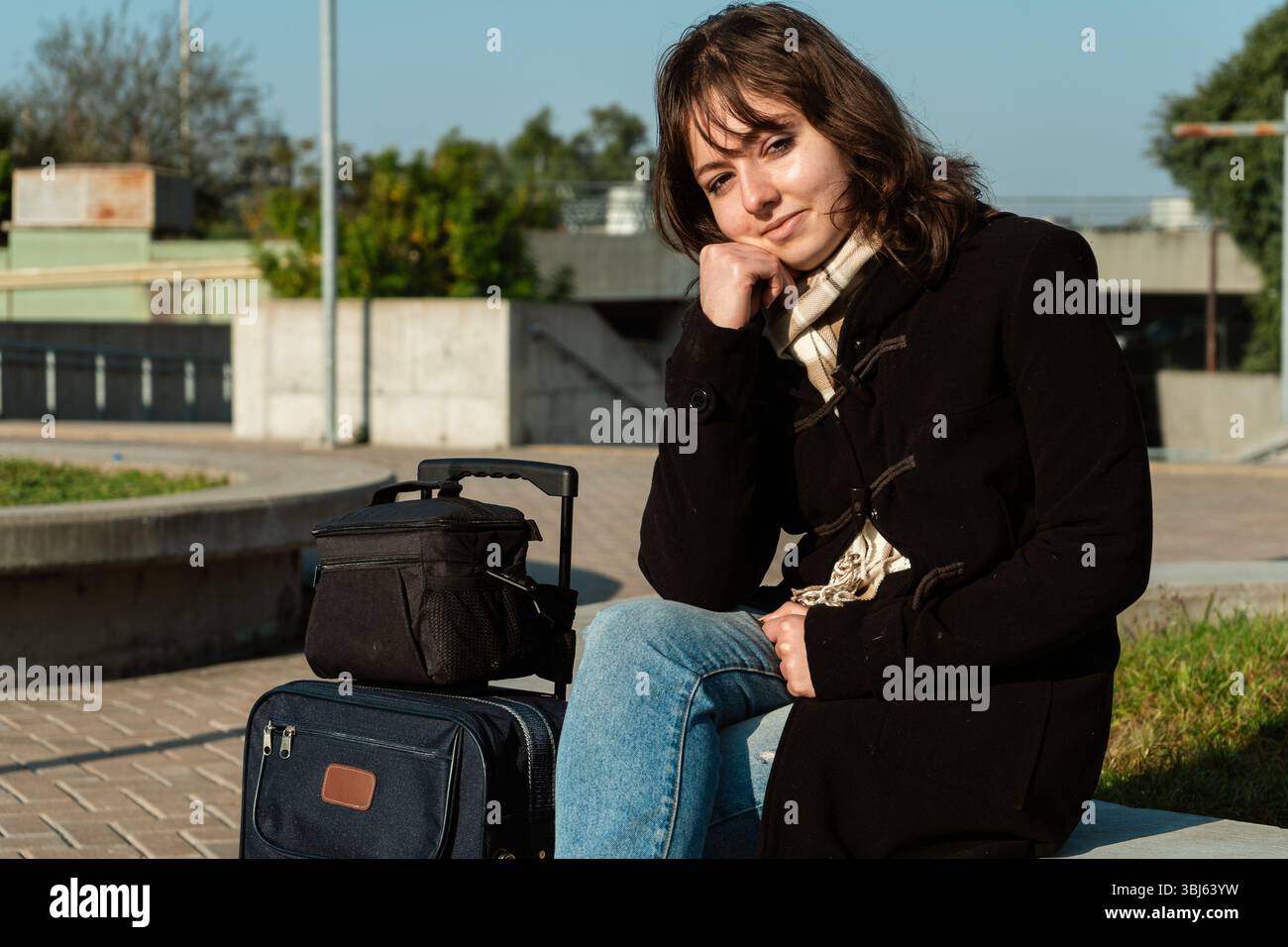 Adult woman with luggage sitting on a concrete bench in a park, calmly ...