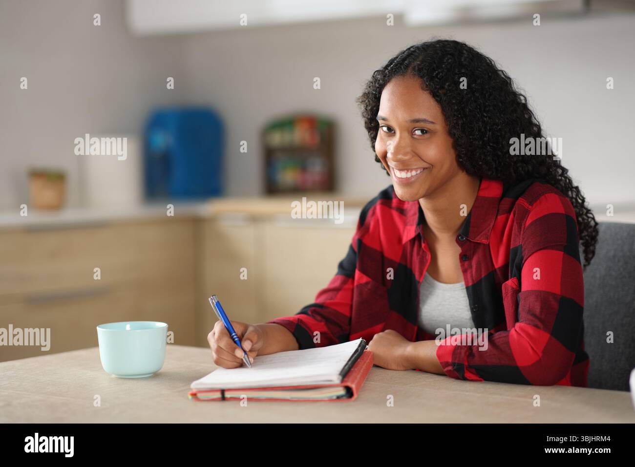 Happy black student taking notes looks at you in the kitchen at home ...