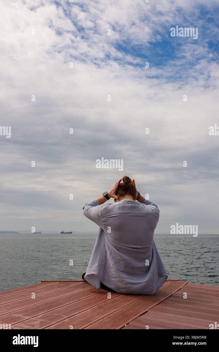 Man sitting cross-legged alone by sea under dramatic cloudy sky, back ...