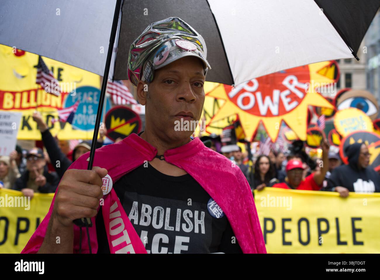 A person at a protest holds a black umbrella. They wear a silver cap ...