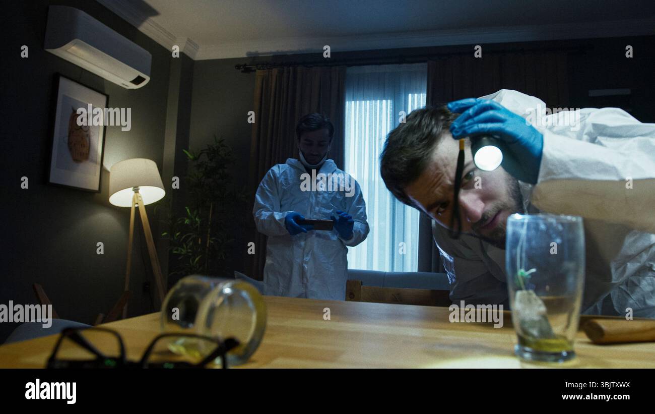 Two forensic experts in protective gear examine clues in a dimly lit ...