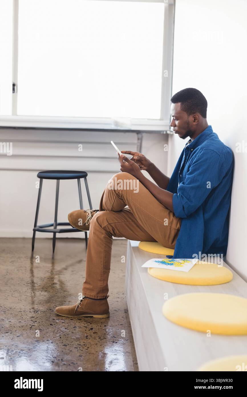 Man sitting on bench in office tapping smartphone by large window with ...