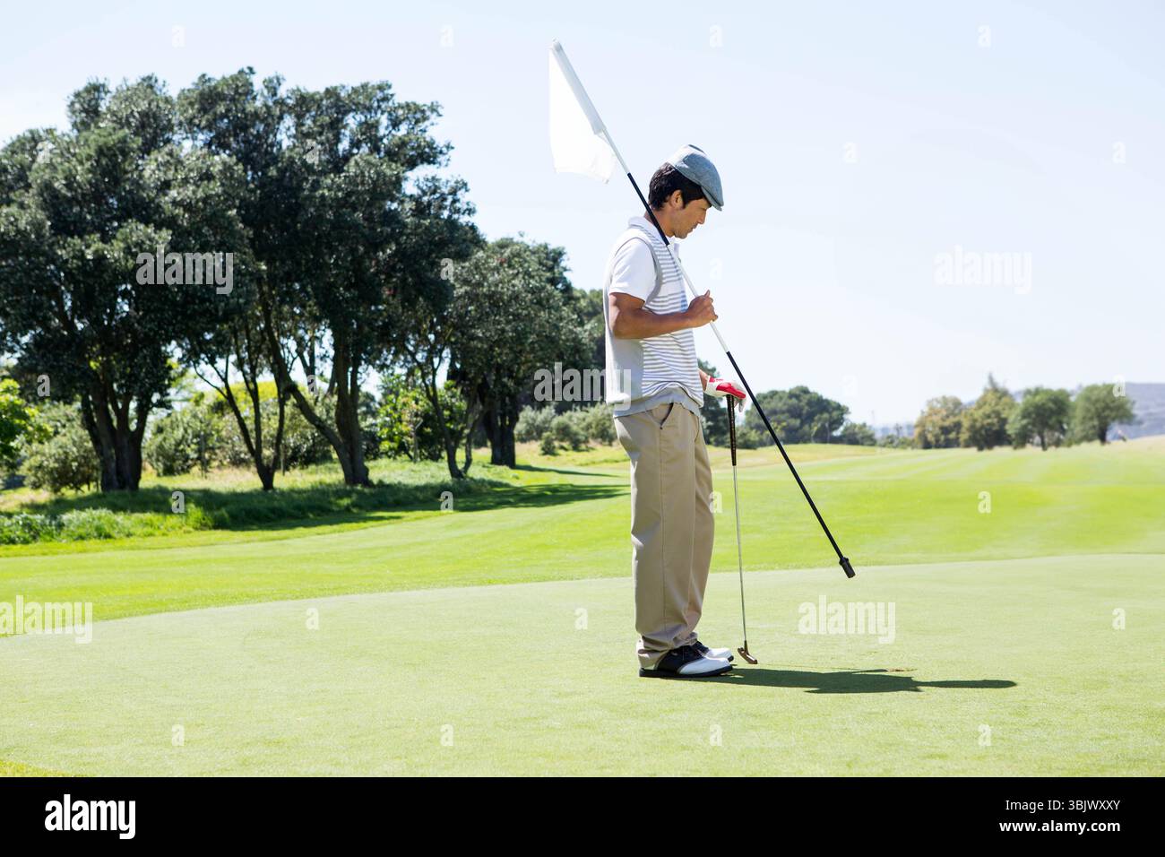 Golfer standing on putting green holding flagstick and putter, gazing ...