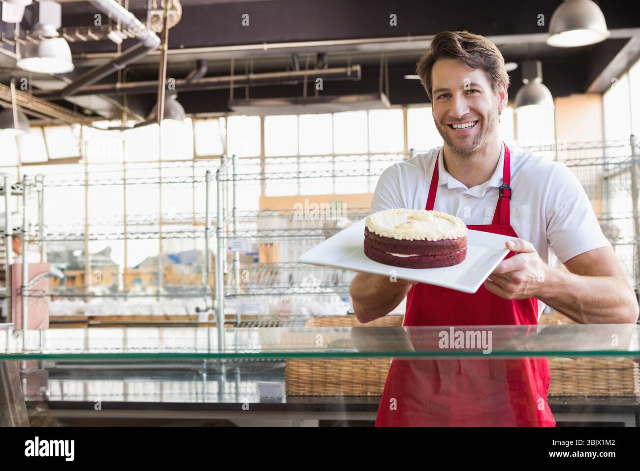Male baker wearing red apron standing at bakery display counter holding ...