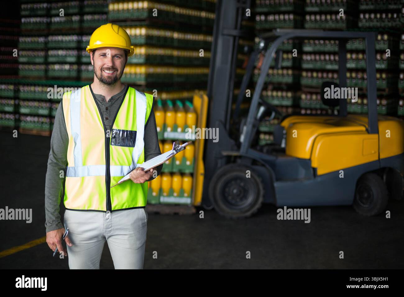 Male warehouse worker wearing helmet, vest holding clipboard near pallets and forklift, copy ...