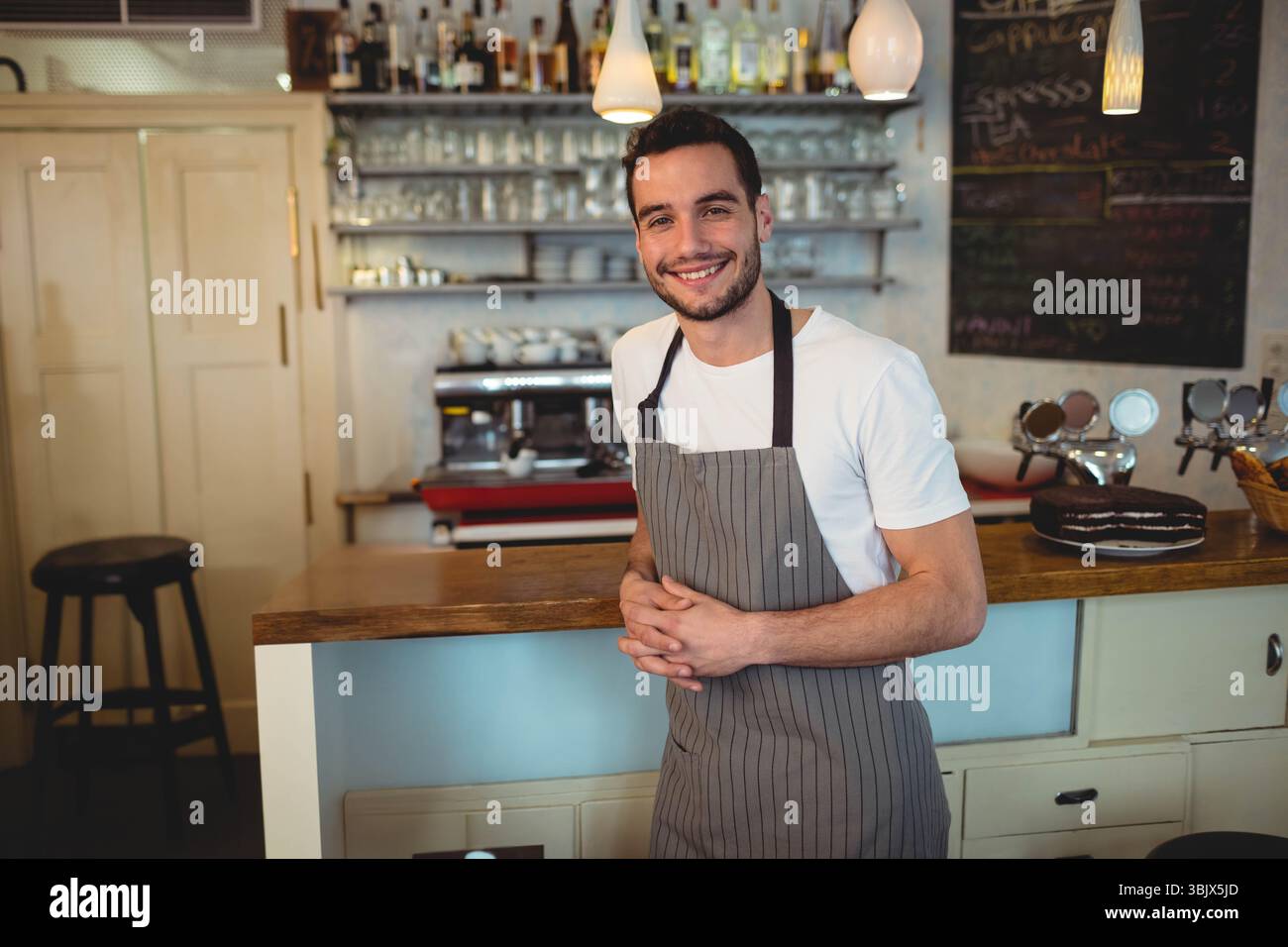 Male cafe worker wearing striped apron standing and smiling behind ...