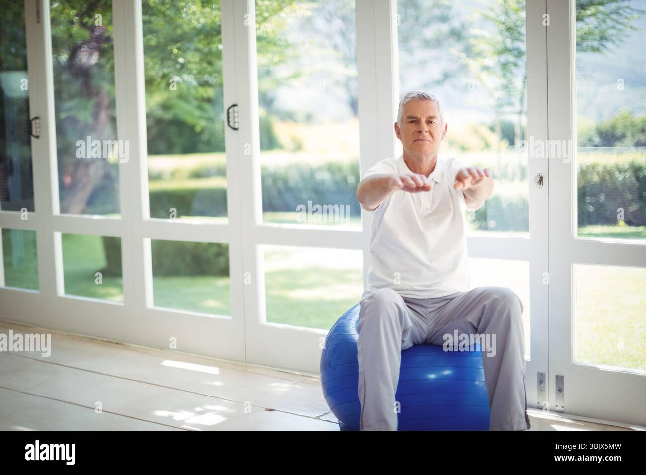 Senior man balancing on blue stability ball in home gym by white-framed ...