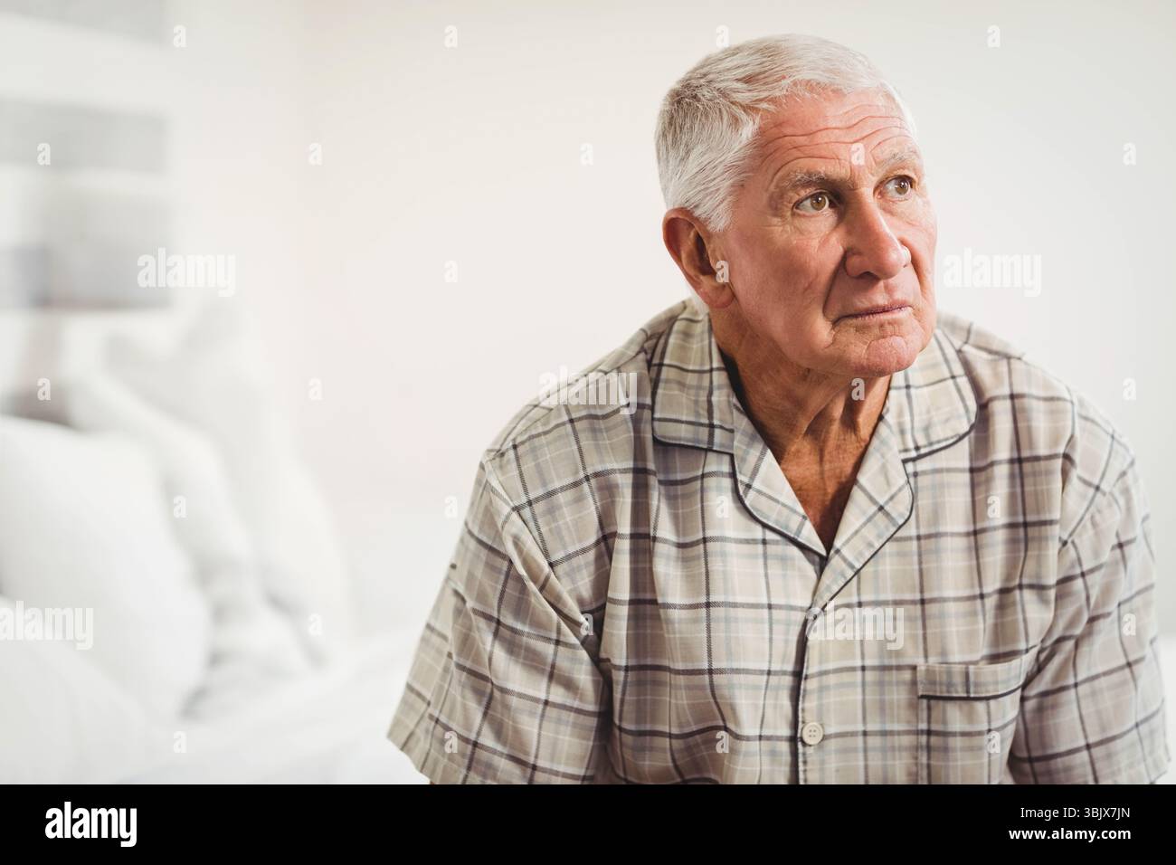 Senior man sitting on bed edge, wearing plaid pajama top in softly lit ...