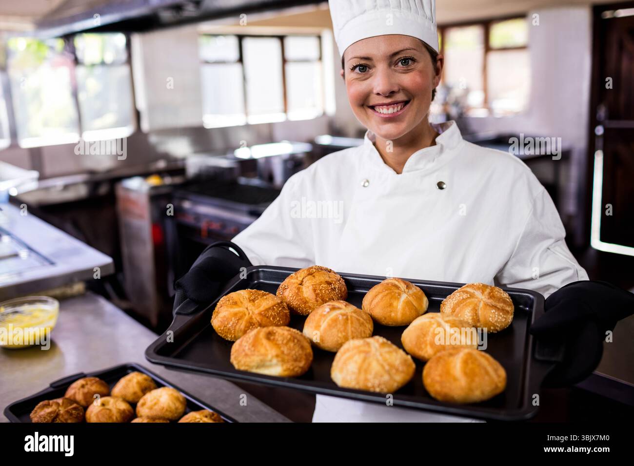 Female chef holding baking tray with sesame-topped bread rolls in commercial kitchen, copy space ...