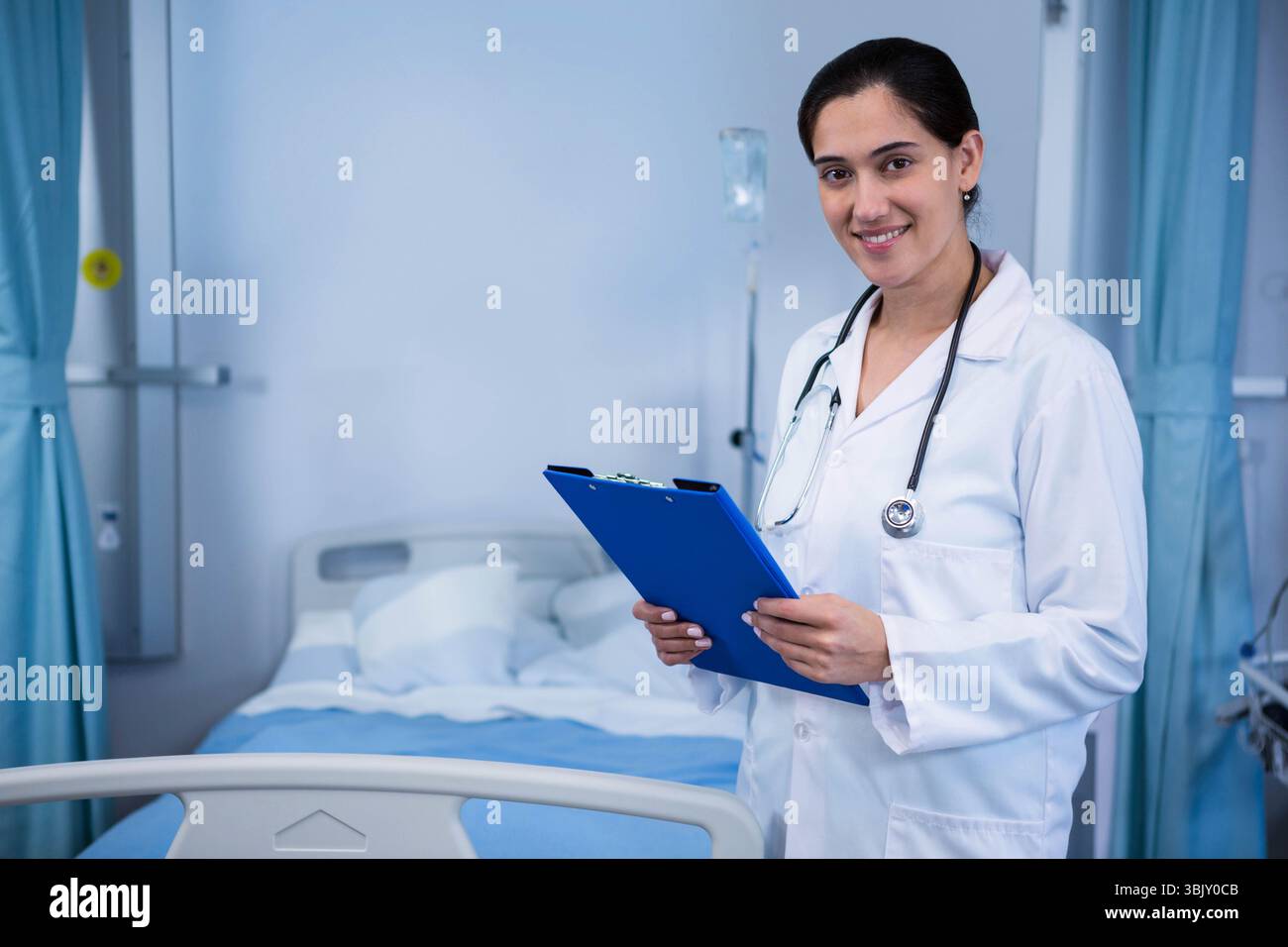 Smiling Indian doctor holding clipboard, wearing stethoscope at bedside ...