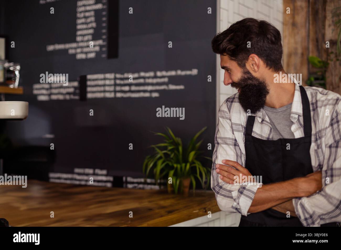 Bearded barista leaning on wooden counter in cafe, with dark menu board ...