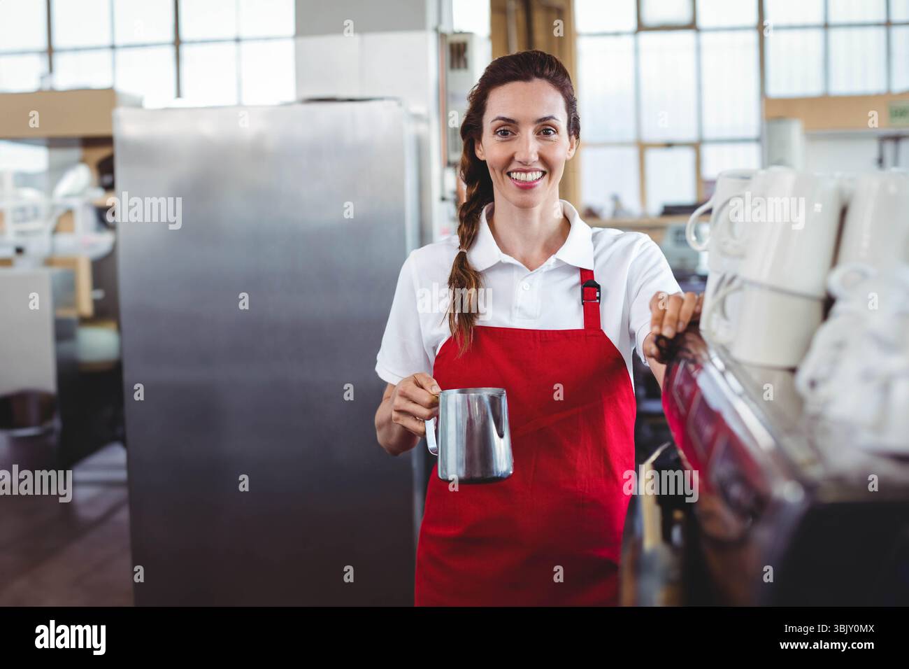Smiling female barista wearing red apron holding milk pitcher at ...